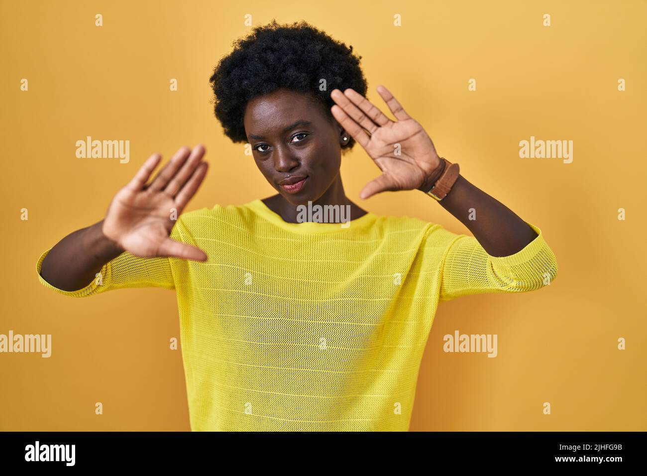African young woman standing over yellow studio doing frame using hands ...