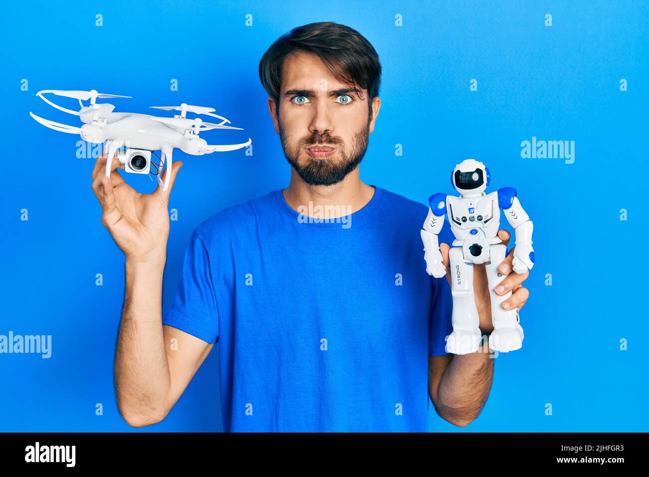 Young hispanic man holding drone and robot toy puffing cheeks with ...