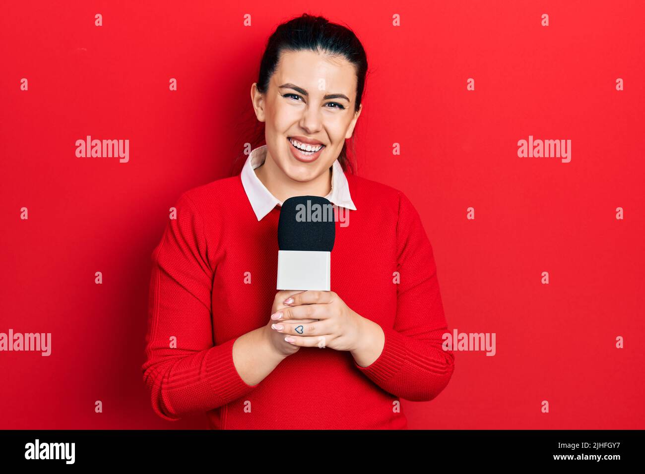 Young hispanic woman holding reporter microphone looking positive and ...