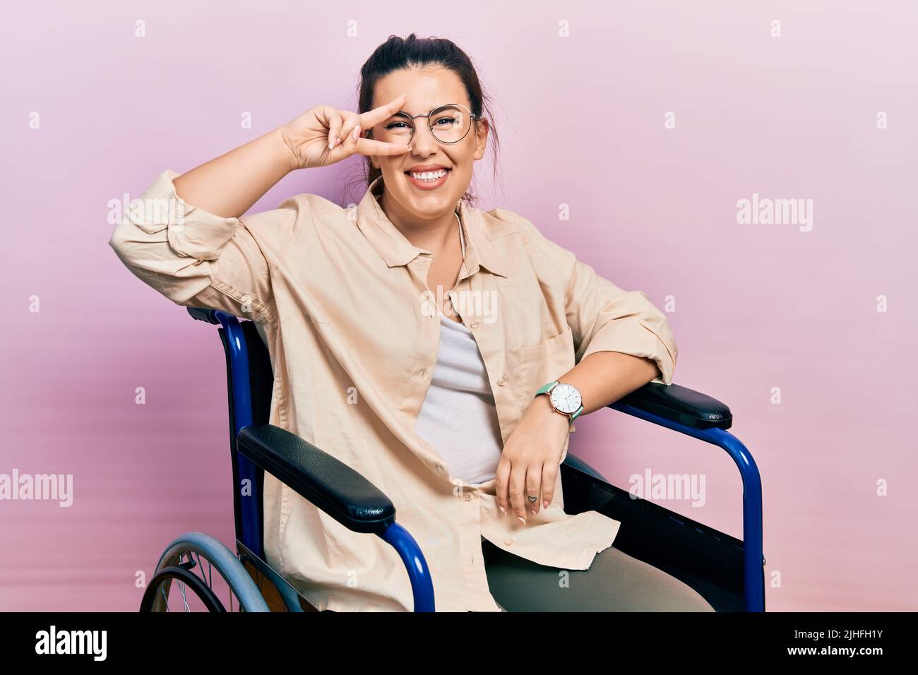 Young hispanic woman sitting on wheelchair doing peace symbol with ...