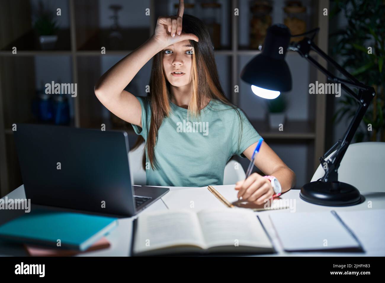 Teenager girl doing homework at home late at night making fun of people with fingers on forehead ...