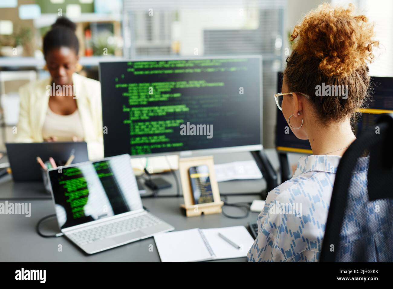 Rear view of young female programmer sitting at her workplace in front of monitors and working ...