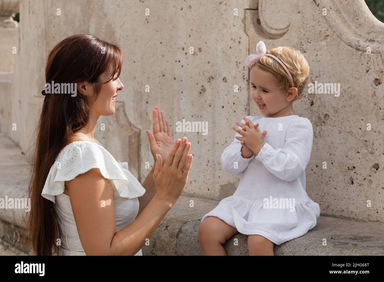 Brunette woman playing patty cake game with baby girl on stone bench in ...