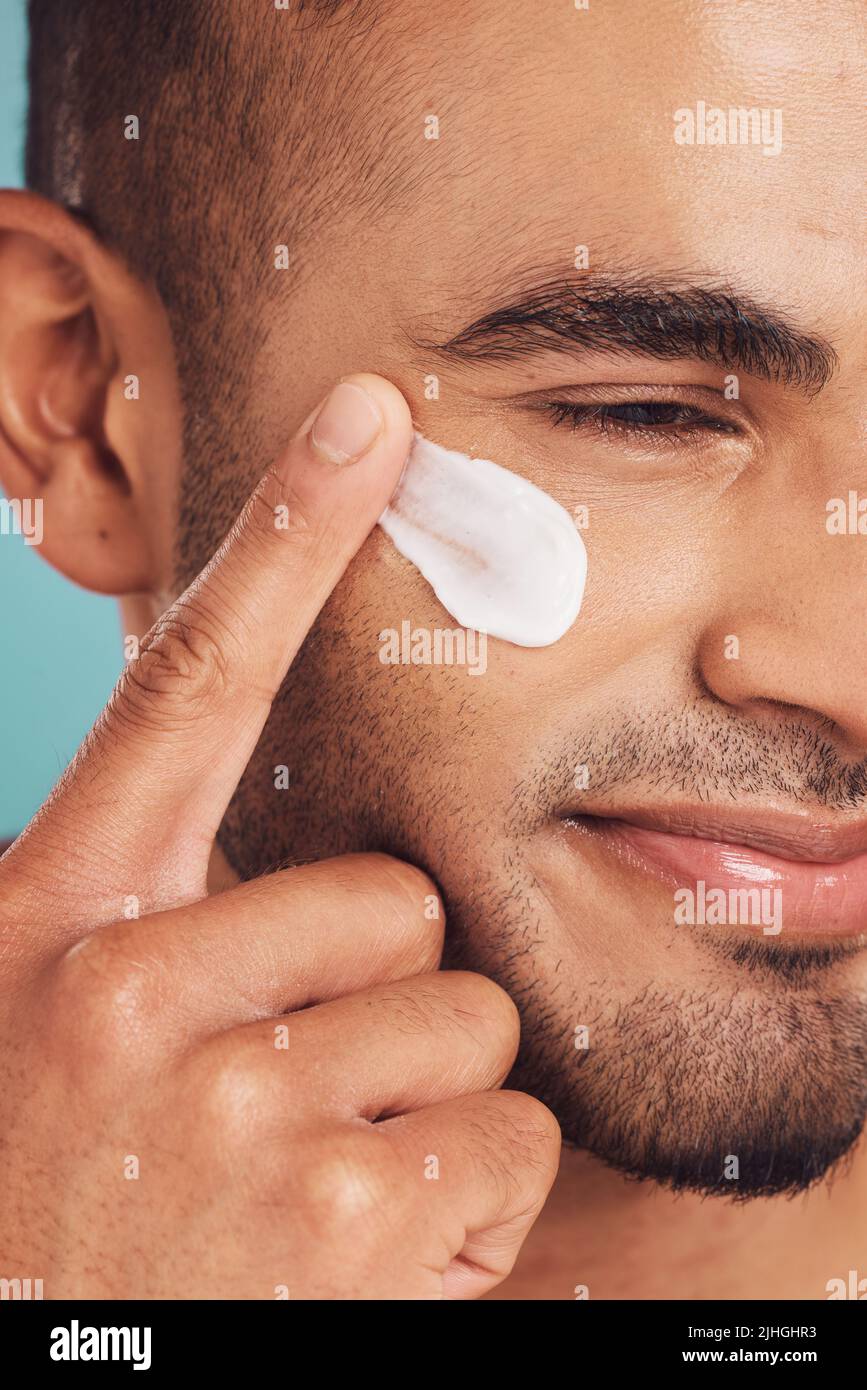 Closeup of one young indian man applying moisturiser lotion to his face while grooming against a ...