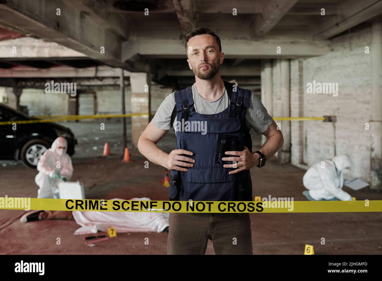 Young serious policeman in bulletproof vest standing behind crime scene ...