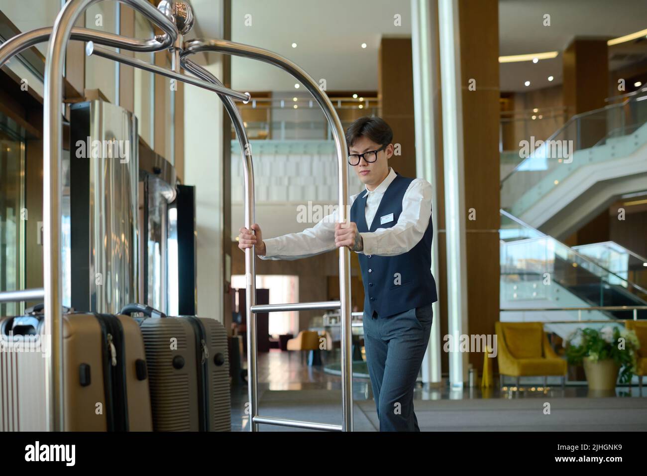 Young bellboy in uniform pushing cart with suitcases of hotel clients ...