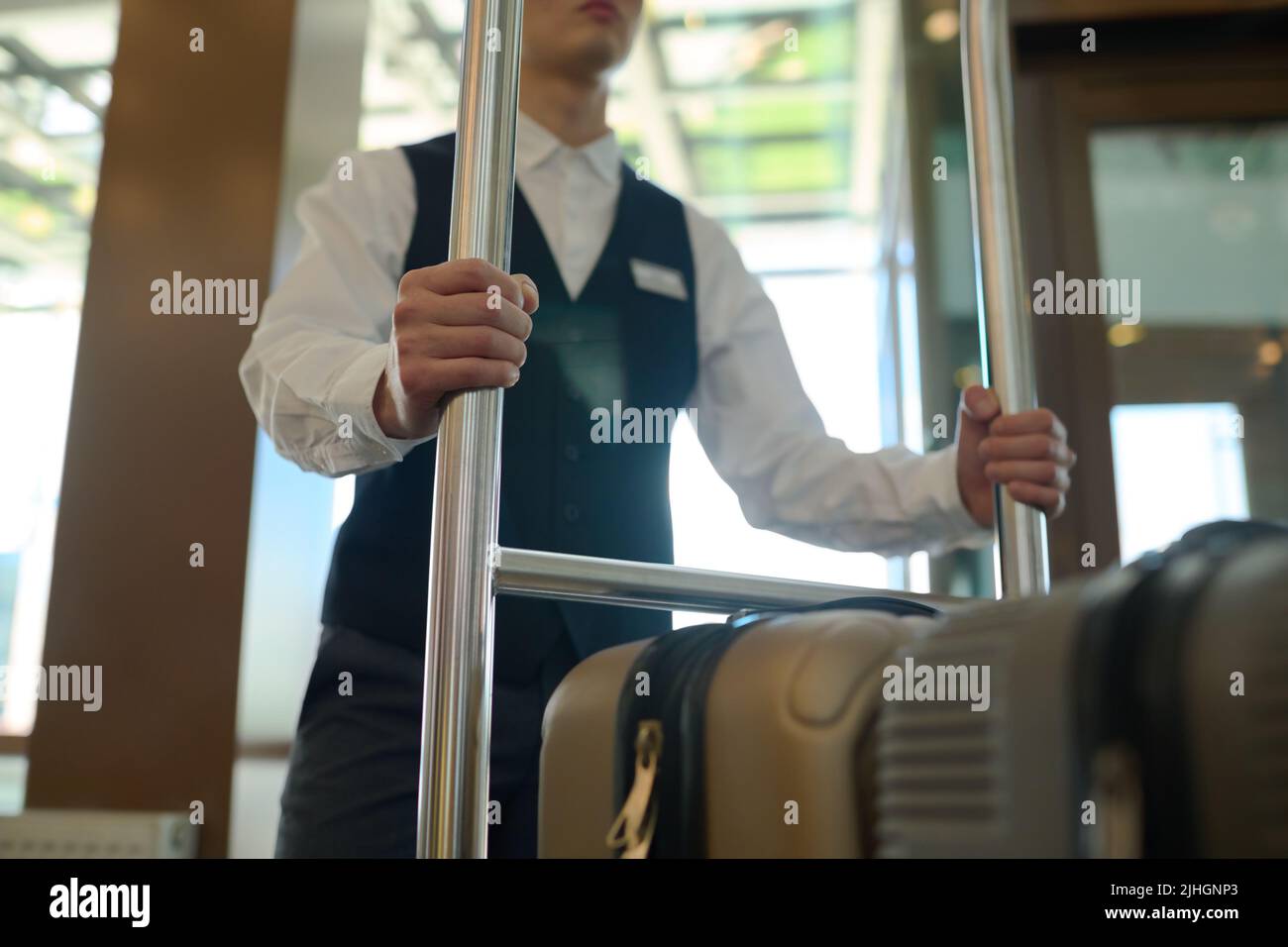 Cropped shot of young bellboy in uniform holding by metallic handles of ...