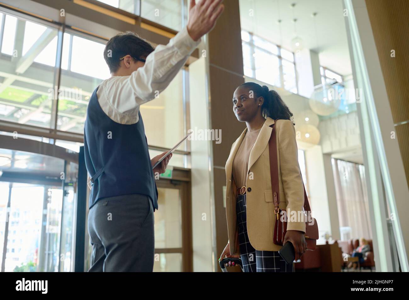 Young owner of hotel meeting one of guests in lounge and pointing right ...