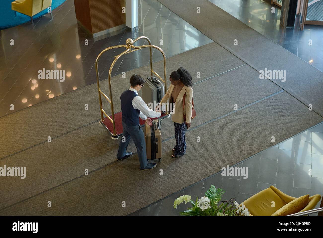 Above angle of young female traveler passing her suitcase to bellboy ...