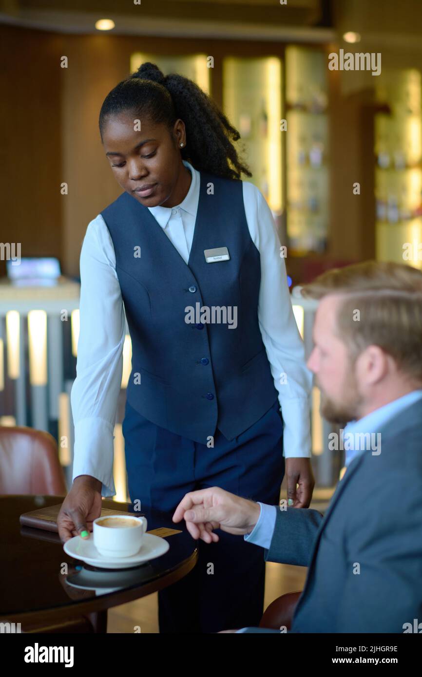 Young African American female manager of hotel in uniform bringing cup ...