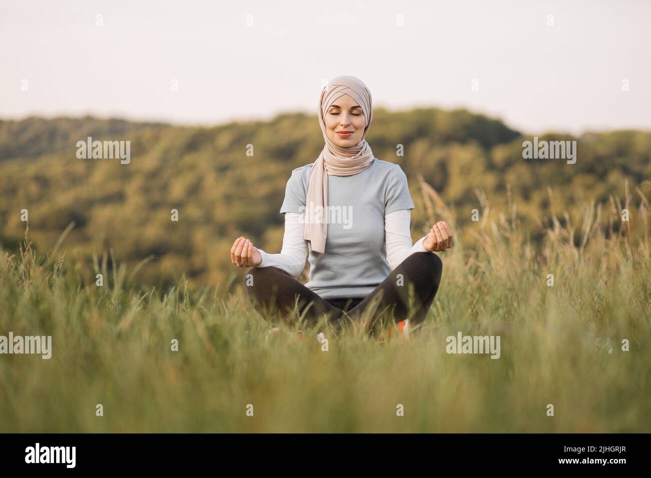 Peaceful young lady in hijab meditating with closed eyes in the park ...