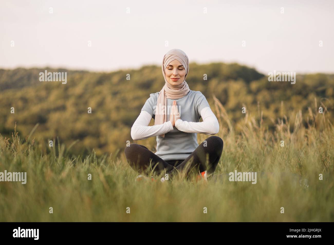 Peaceful young lady in hijab meditating with closed eyes and namaste ...