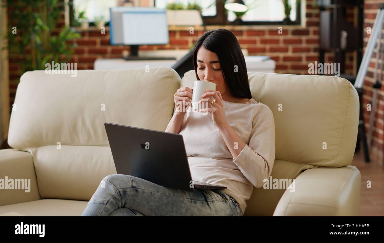 Happy asian woman sipping beverage while working remotely on laptop at ...