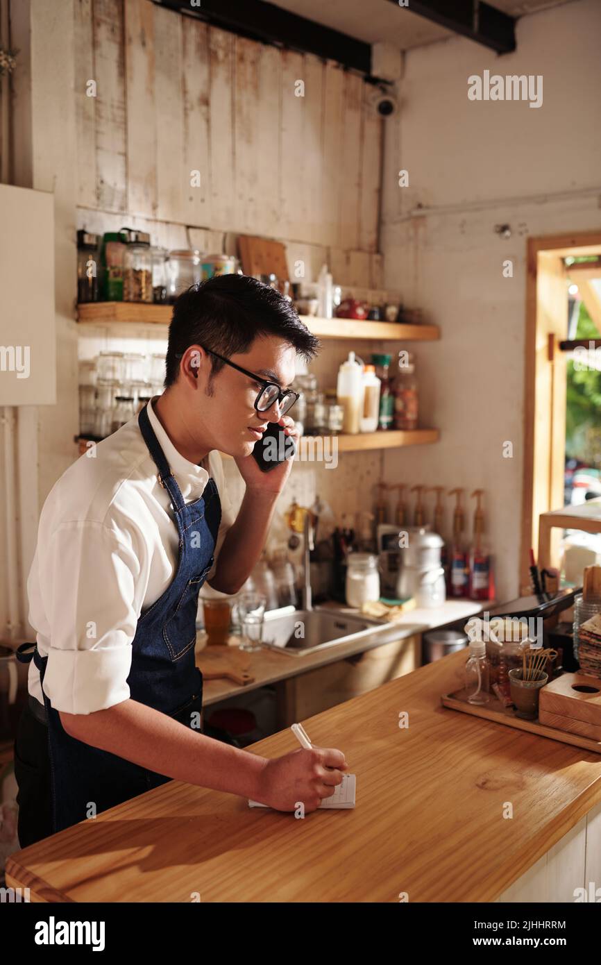 Young barista accepting order from customer in phone and writing it on ...
