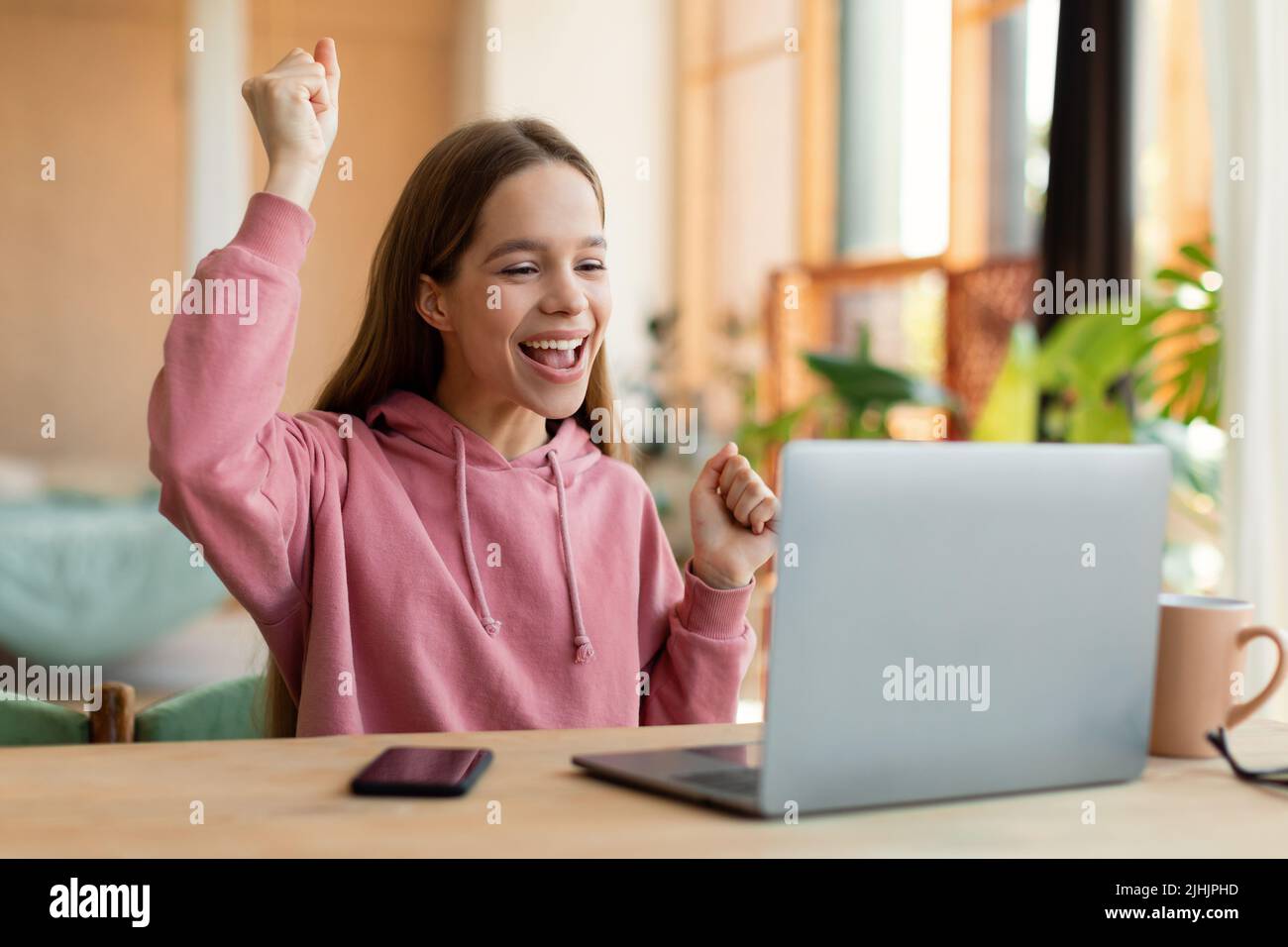Overjoyed teen girl sitting at desk with laptop and making YES gesture ...