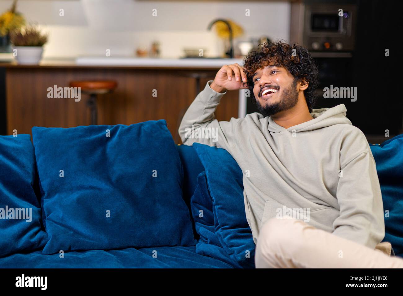 Ethnic unshaven man sitting on the blue cozy sofa and smiling, while ...