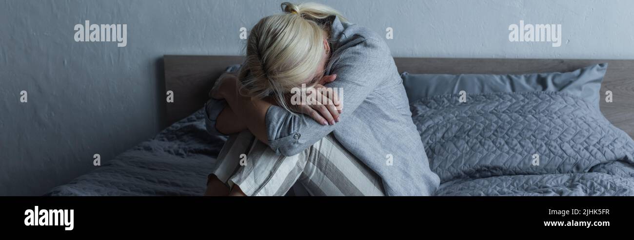 blonde woman covering face and hugging knees on bed while feeling pain ...