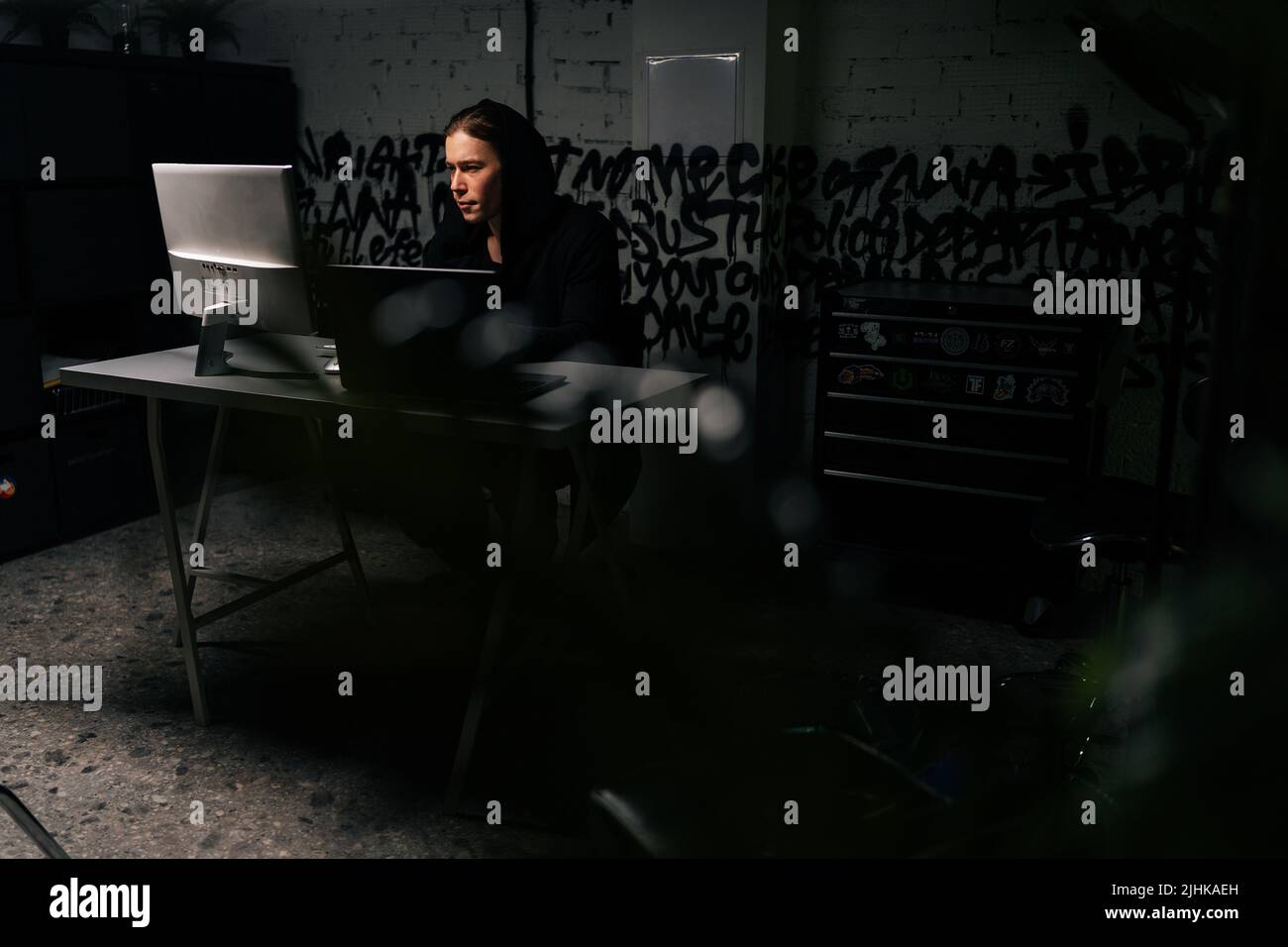 Focused hacker man in black sweatshirt using laptop and desktop computer to hack network system ...