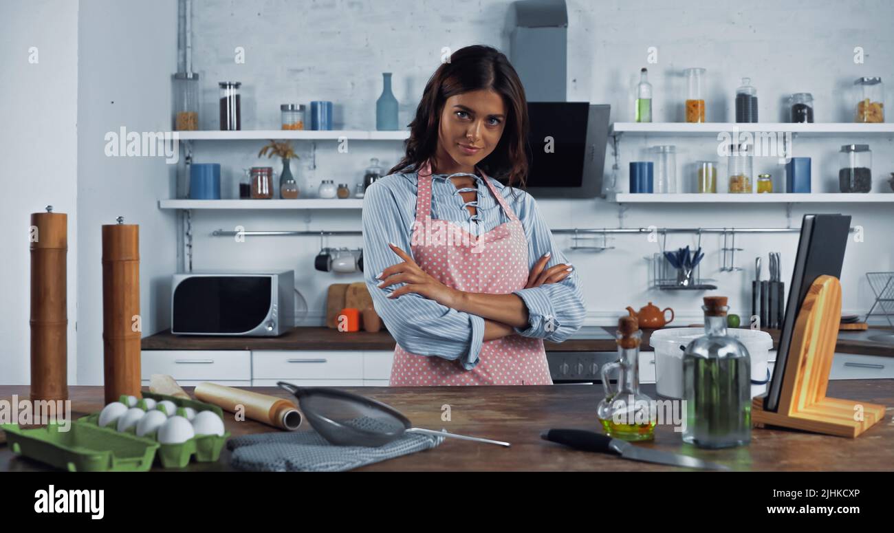 brunette woman standing with crossed arms near ingredients and cooking ...