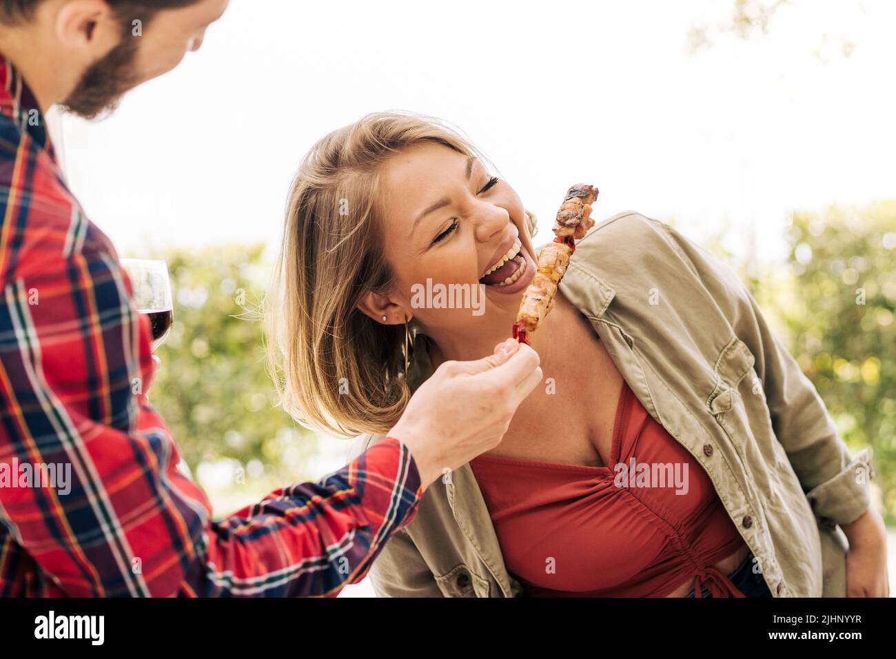 A young curvy woman laughs as she eats skewers of meat from her friend ...