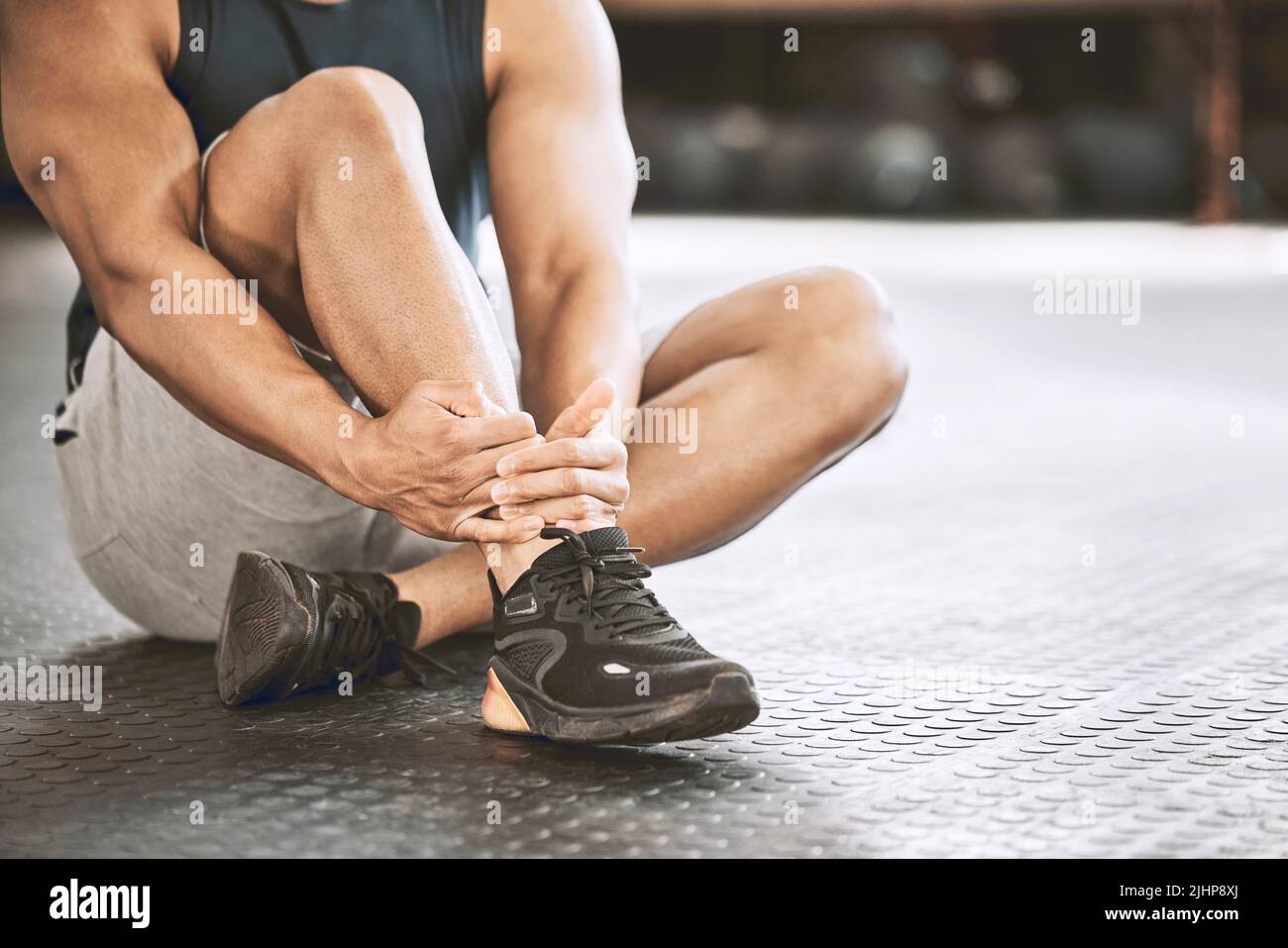 Athlete touching his sprained ankle. Closeup of hands of bodybuilder sitting on the gym floor ...