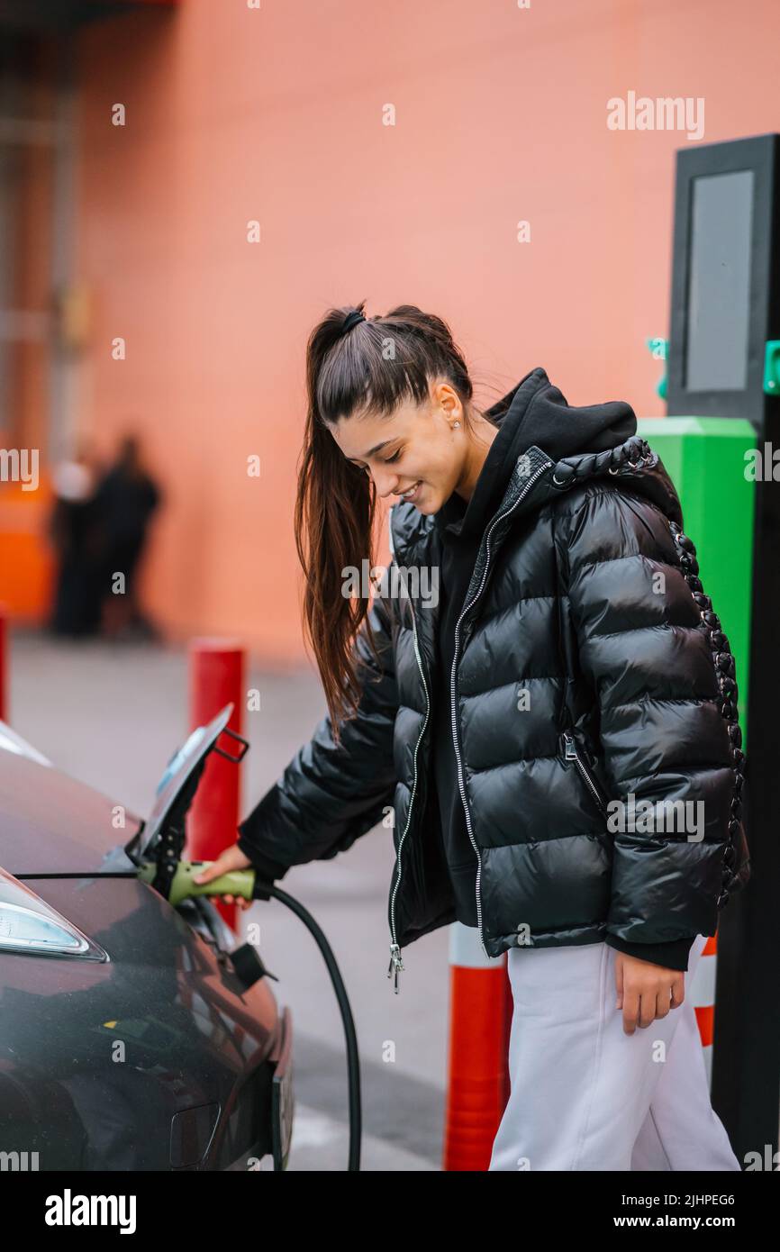 Young beautiful woman traveling by electric car having stop at charging ...