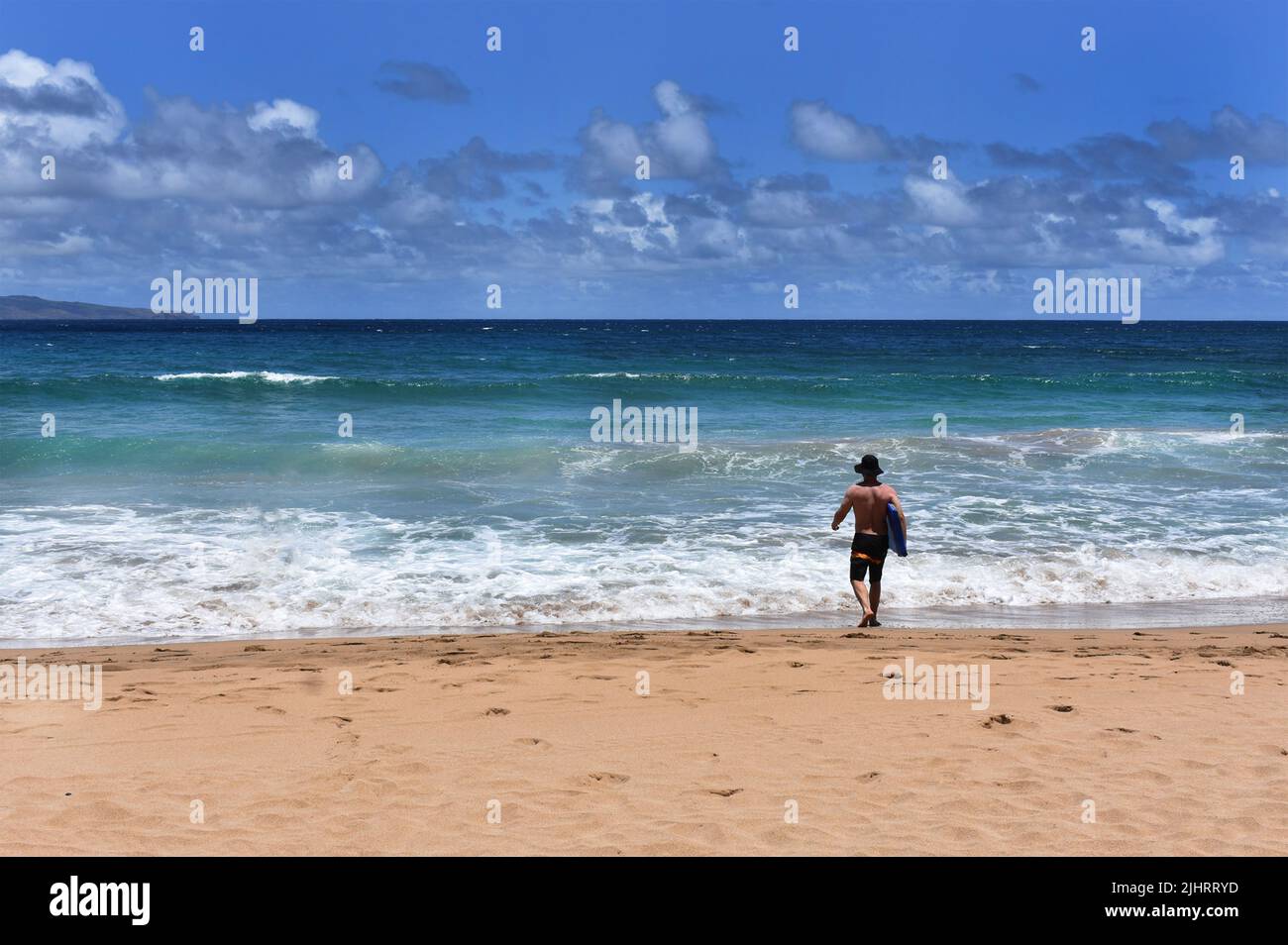 A back view of a man with surfboard on the sandy beach approaching the ...