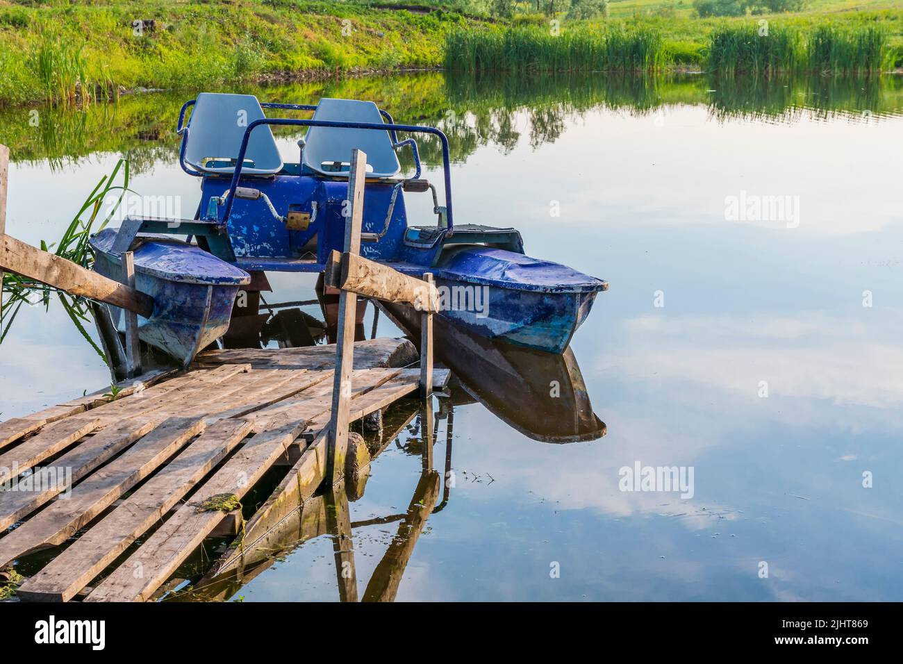 There is old blue catamaran near shore of lake, wooden bridge leads to ...
