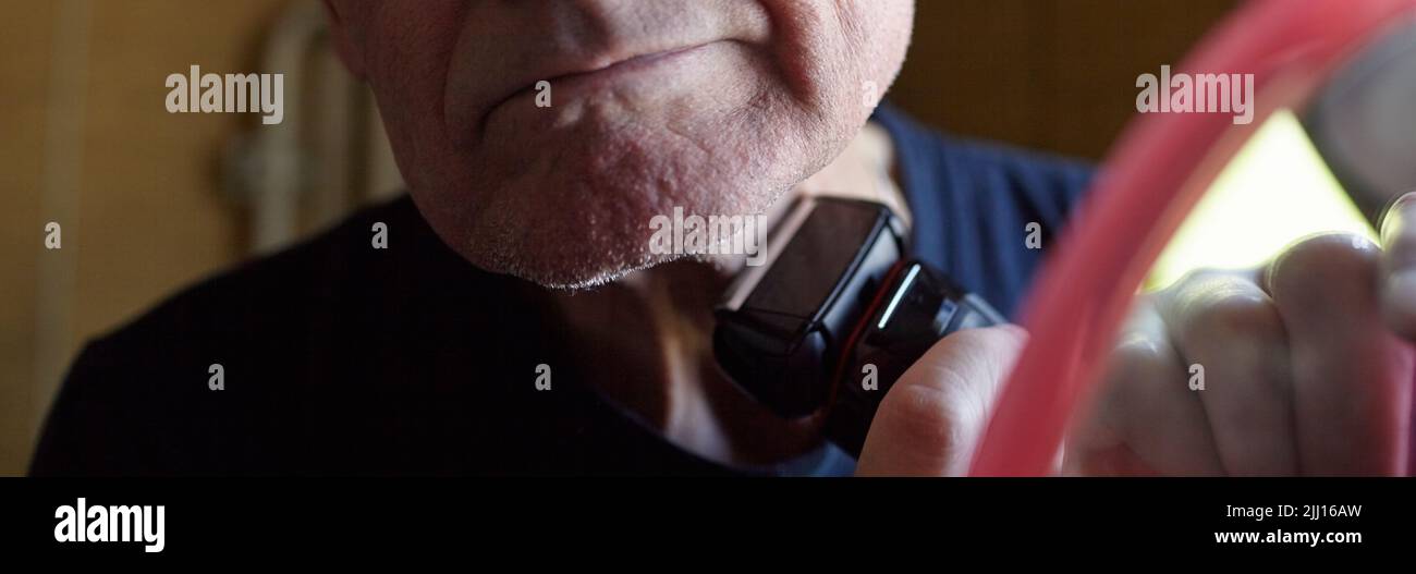 Close up senior man shaving beard with electric razor with small mirror ...