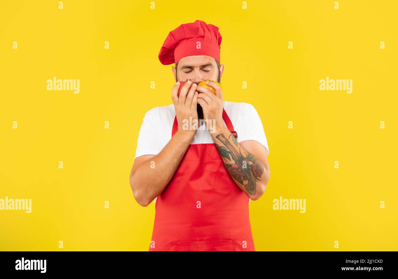 Serious man in cooking apron and toque smelling tomatoes with closed ...