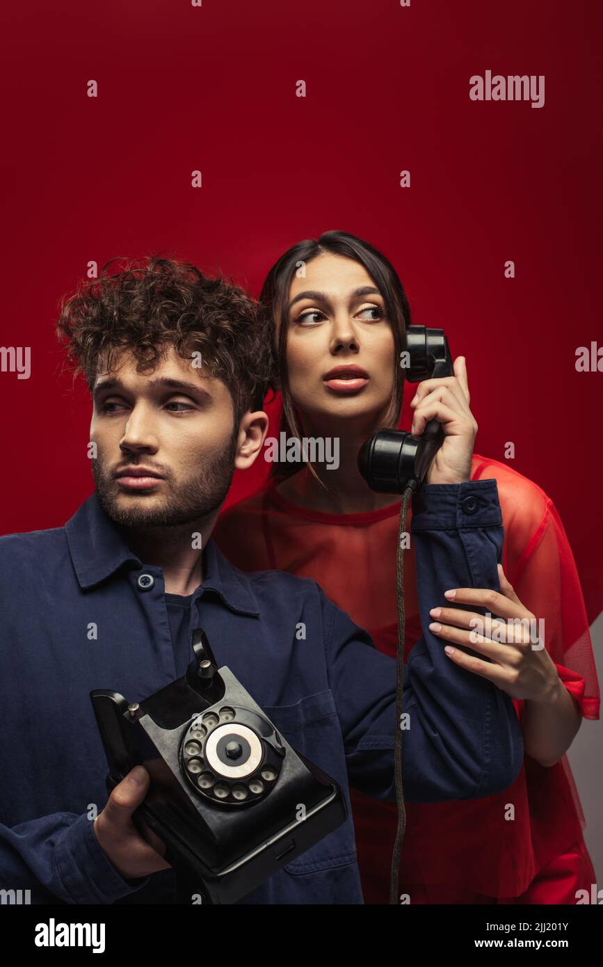 curly man holding handset and retro telephone near young woman on red ...