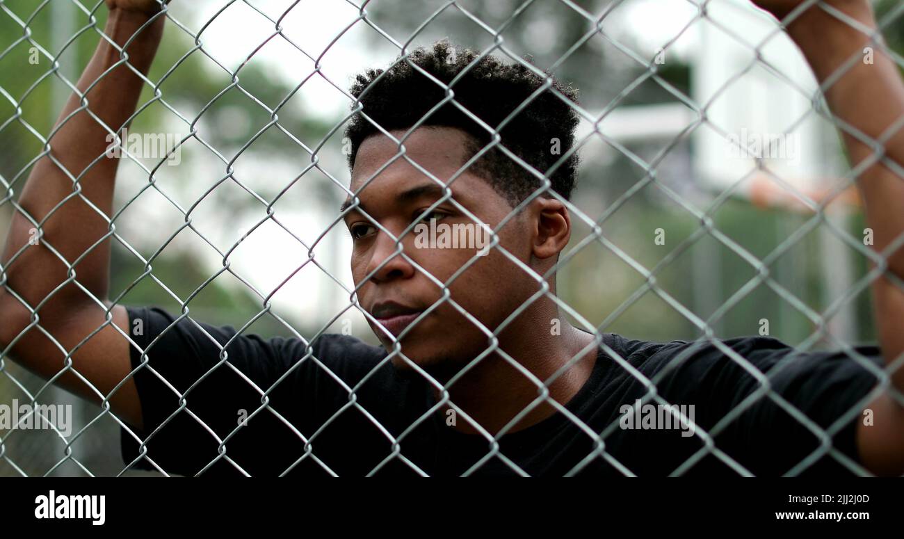 African mixed race man standing behind metal fence. Pensive black guy ...