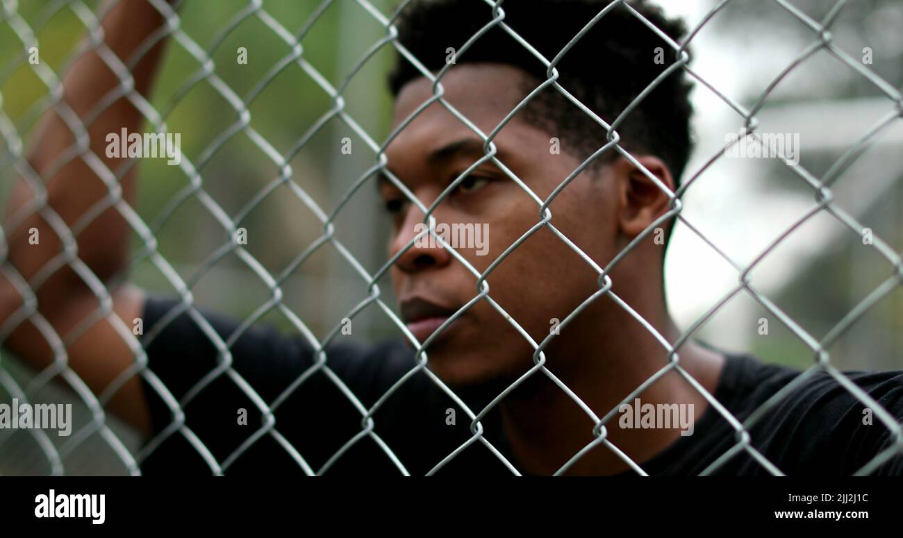 African mixed race man standing behind metal fence. Pensive black guy ...