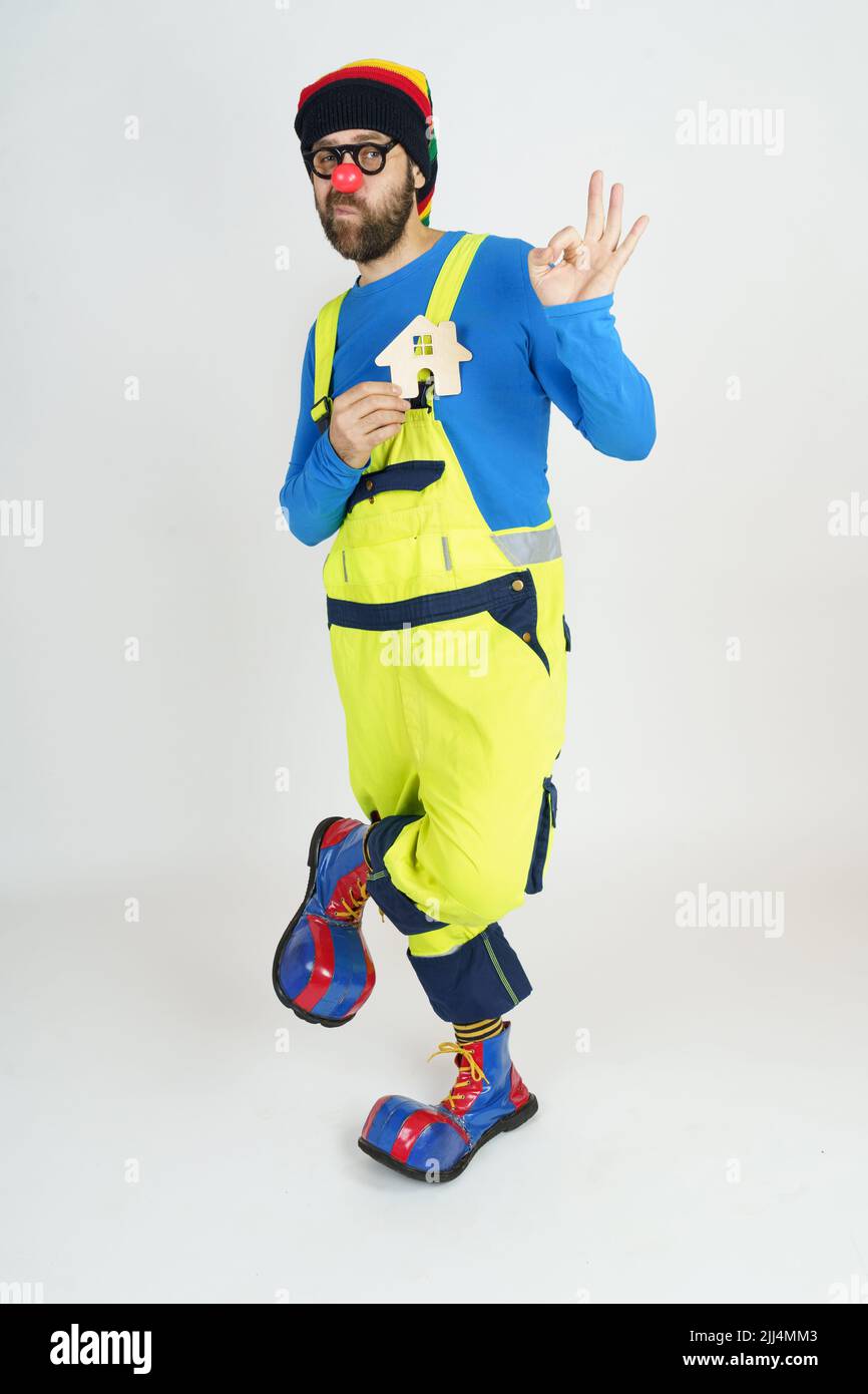 Humor and fun concept. A clown in a bright costume holds a wooden house ...