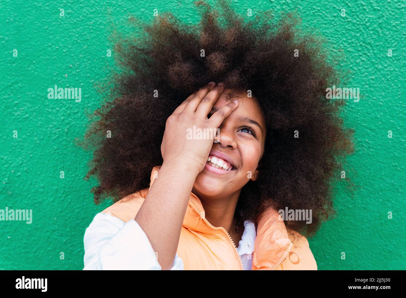 Happy girl with Afro hairstyle covering eye in front of green wall ...