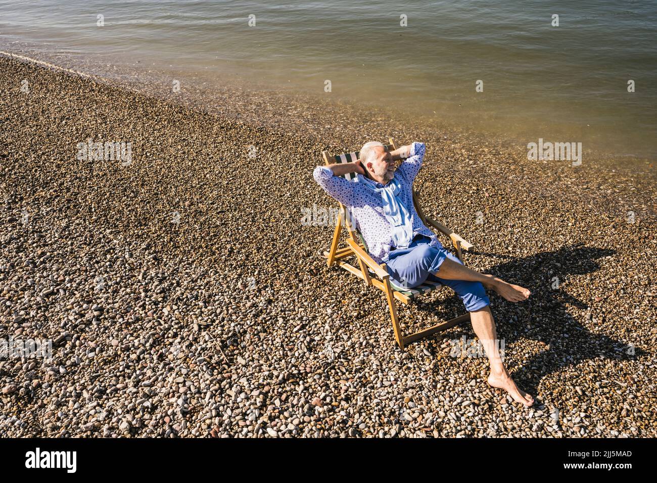 Senior man with hands behind head resting on deck chair at riverbank ...