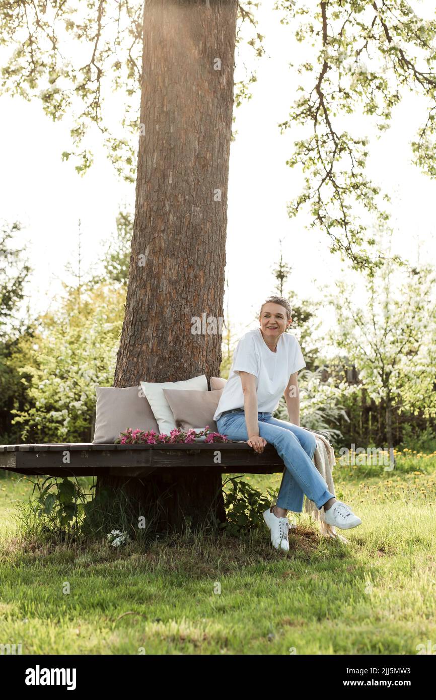 Happy woman sitting on bench around tree trunk in garden Stock Photo ...