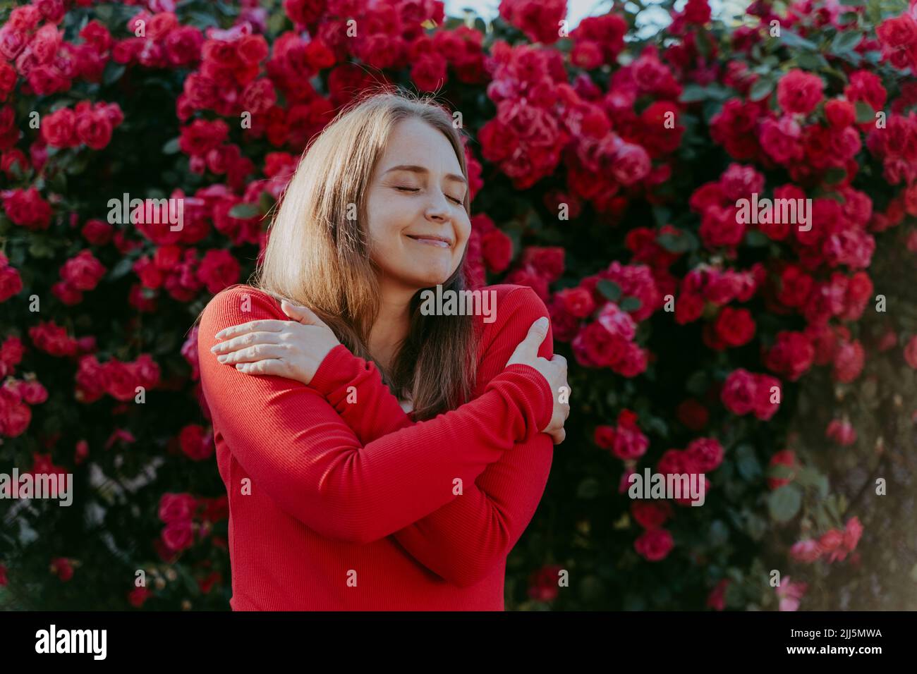 Smiling woman hugging self in front of rose bush Stock Photo - Alamy