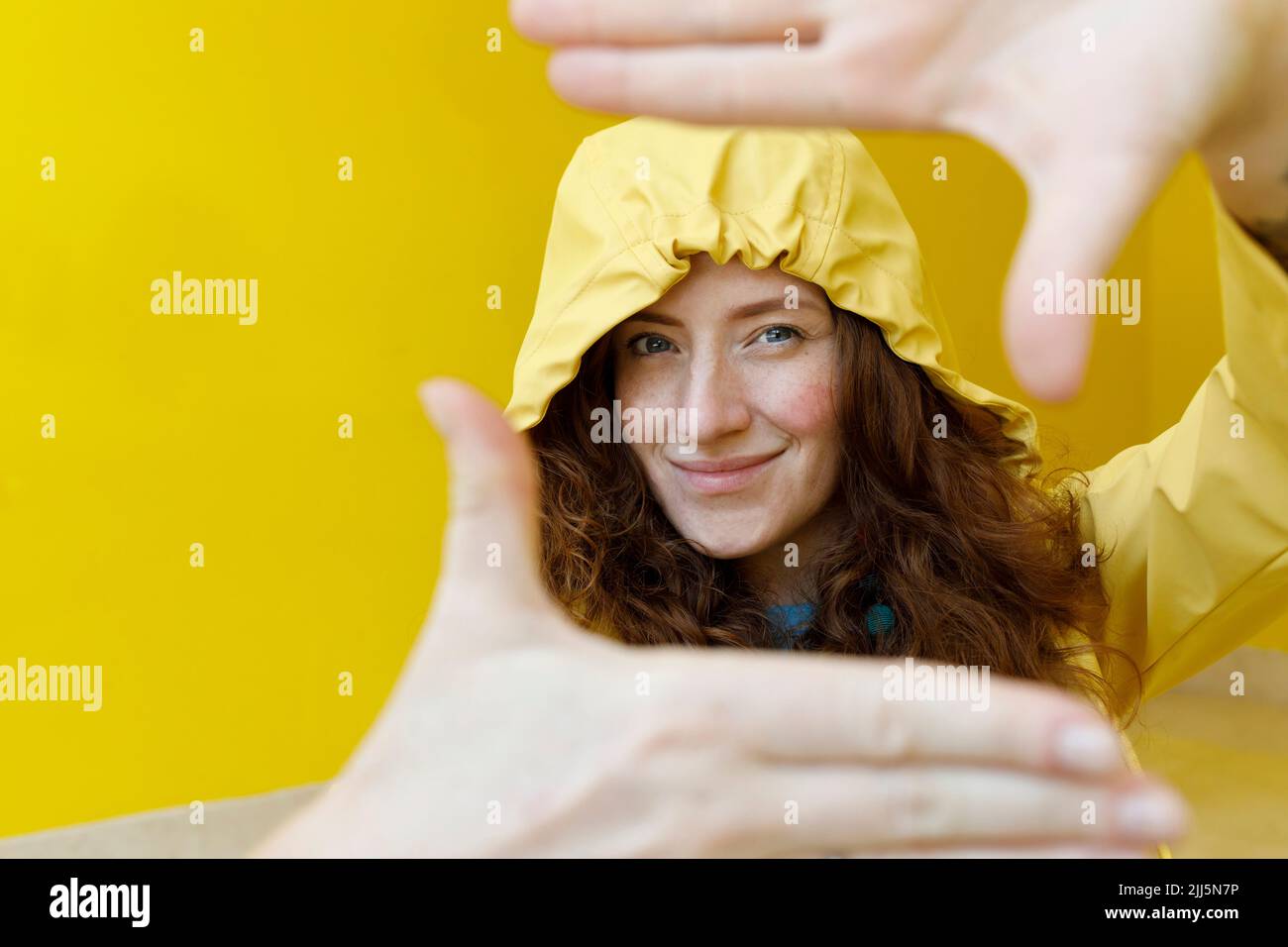 Smiling woman in yellow raincoat making frame with hands in front of ...