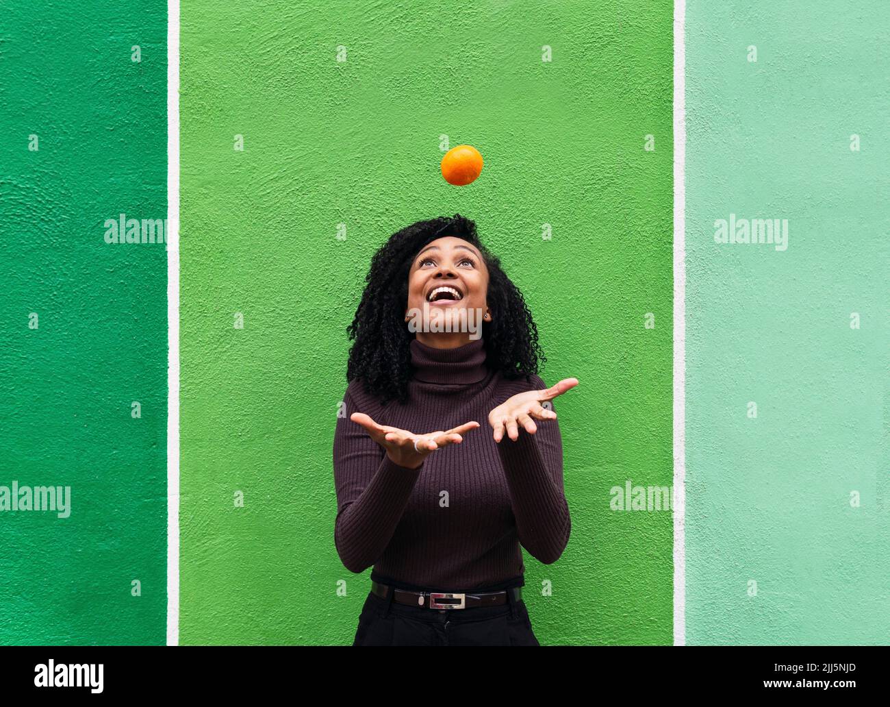 Cheerful woman with mouth open throwing orange in front of green wall ...