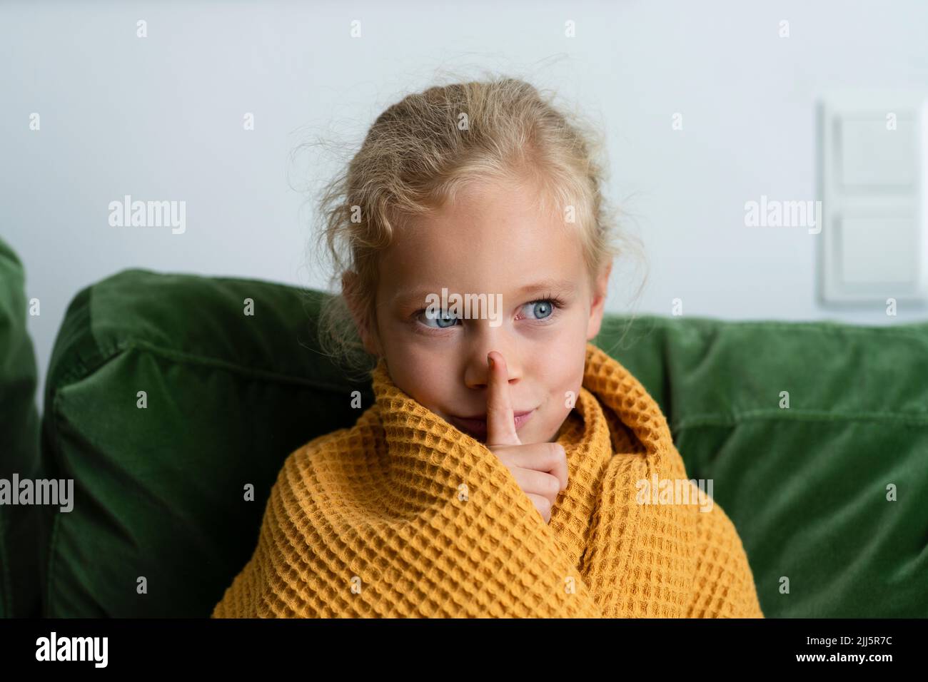Contemplative girl wrapped in blanket making silence gesture at home ...