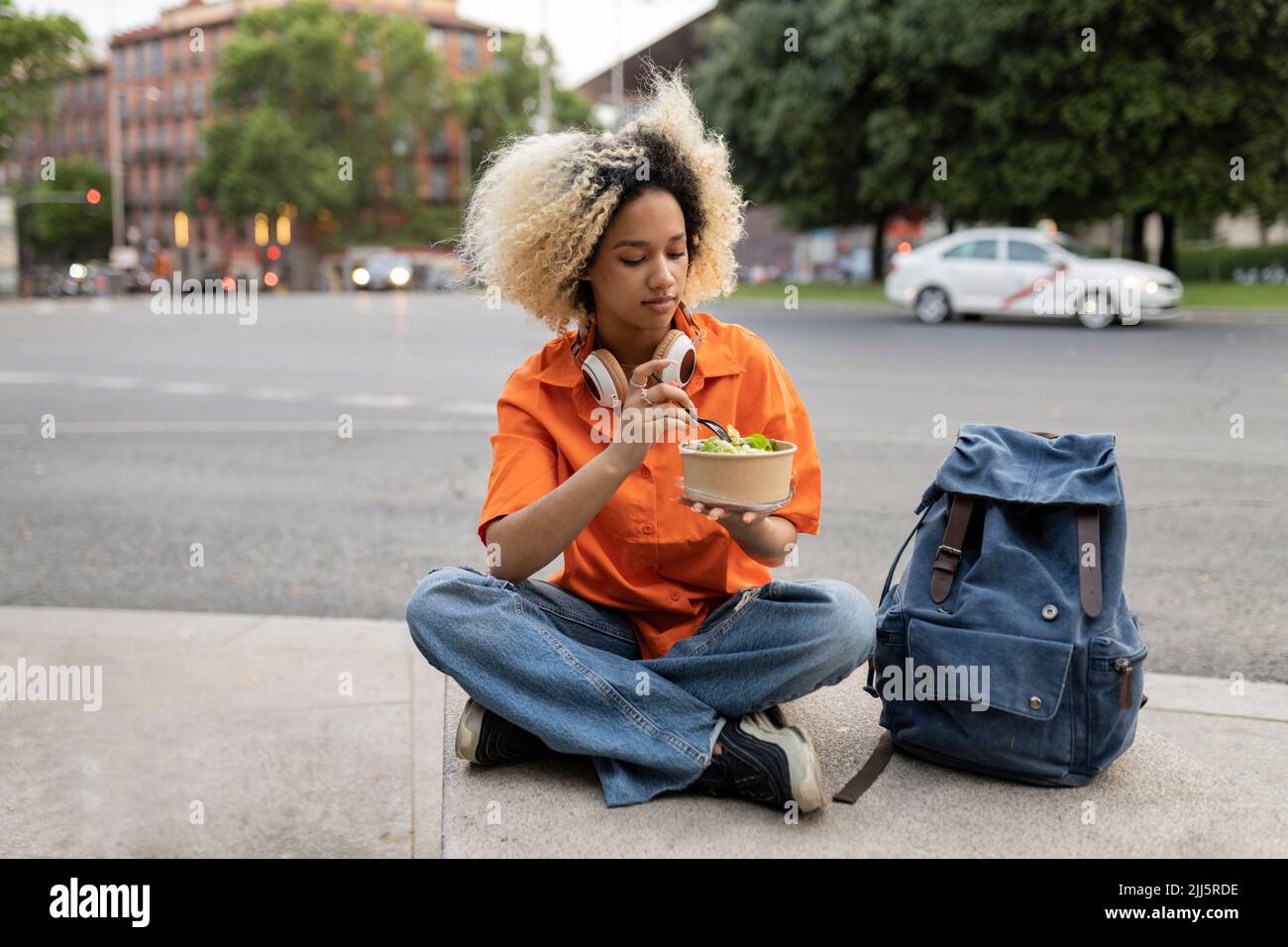 Woman eating lunch sitting cross-legged by backpack on sidewalk Stock ...