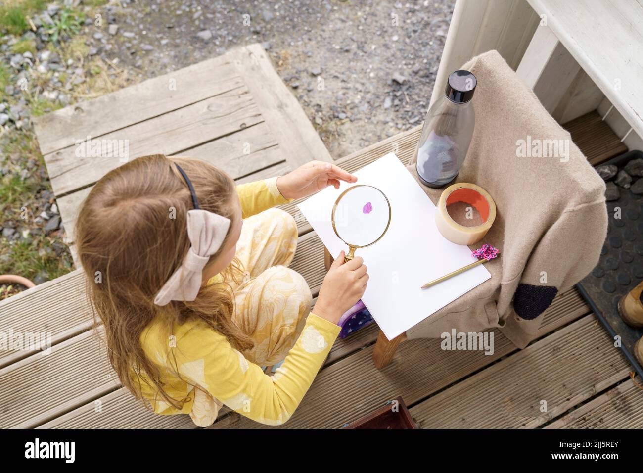 Girl observing flower petal though magnifying glass sitting at porch ...