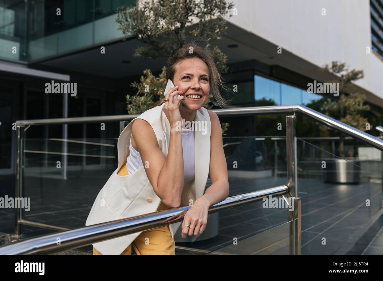Smiling businesswoman talking through smart phone leaning on railing ...