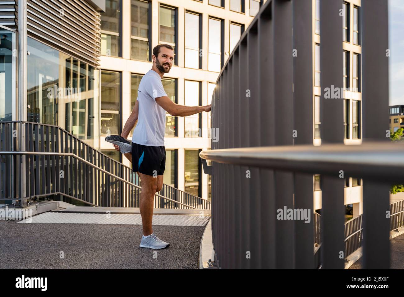 Smiling man stretching leg standing by railing on walkway Stock Photo ...