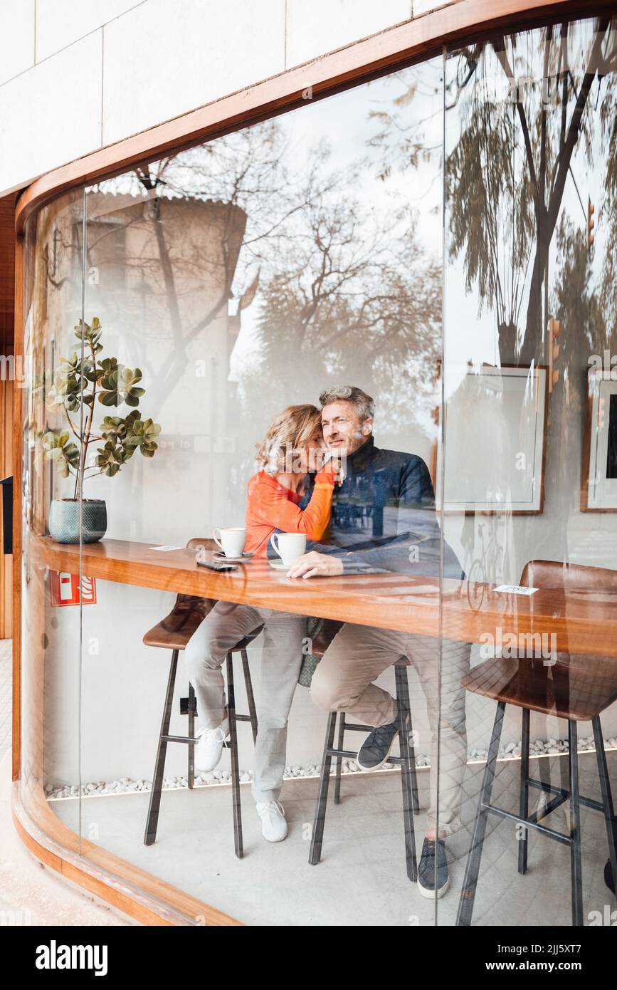 Woman embracing man sitting on chair in cafe seen through window Stock ...