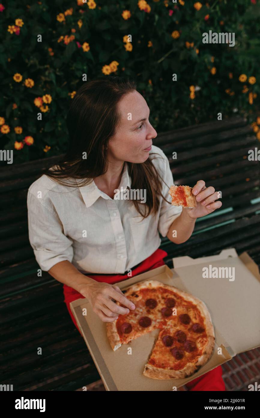 Woman eating slice of pizza sitting on bench Stock Photo - Alamy