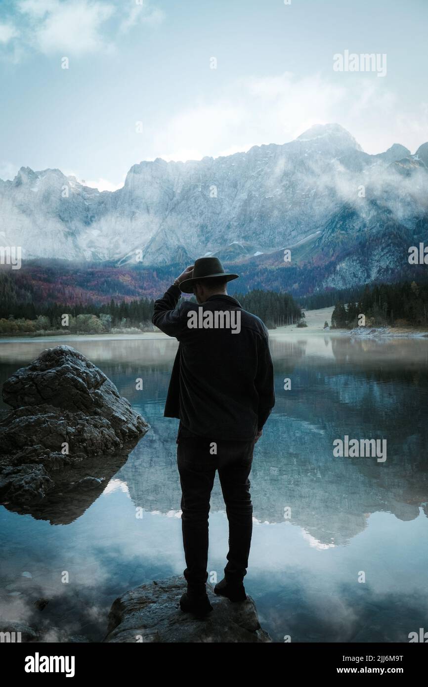 A vertical back view of man with fedora hat standing on rock near lake ...
