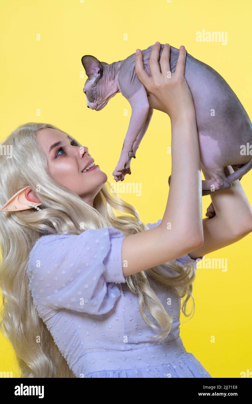 Young blonde woman cosplay elf in blue summer dress holds Sphinx kitten ...