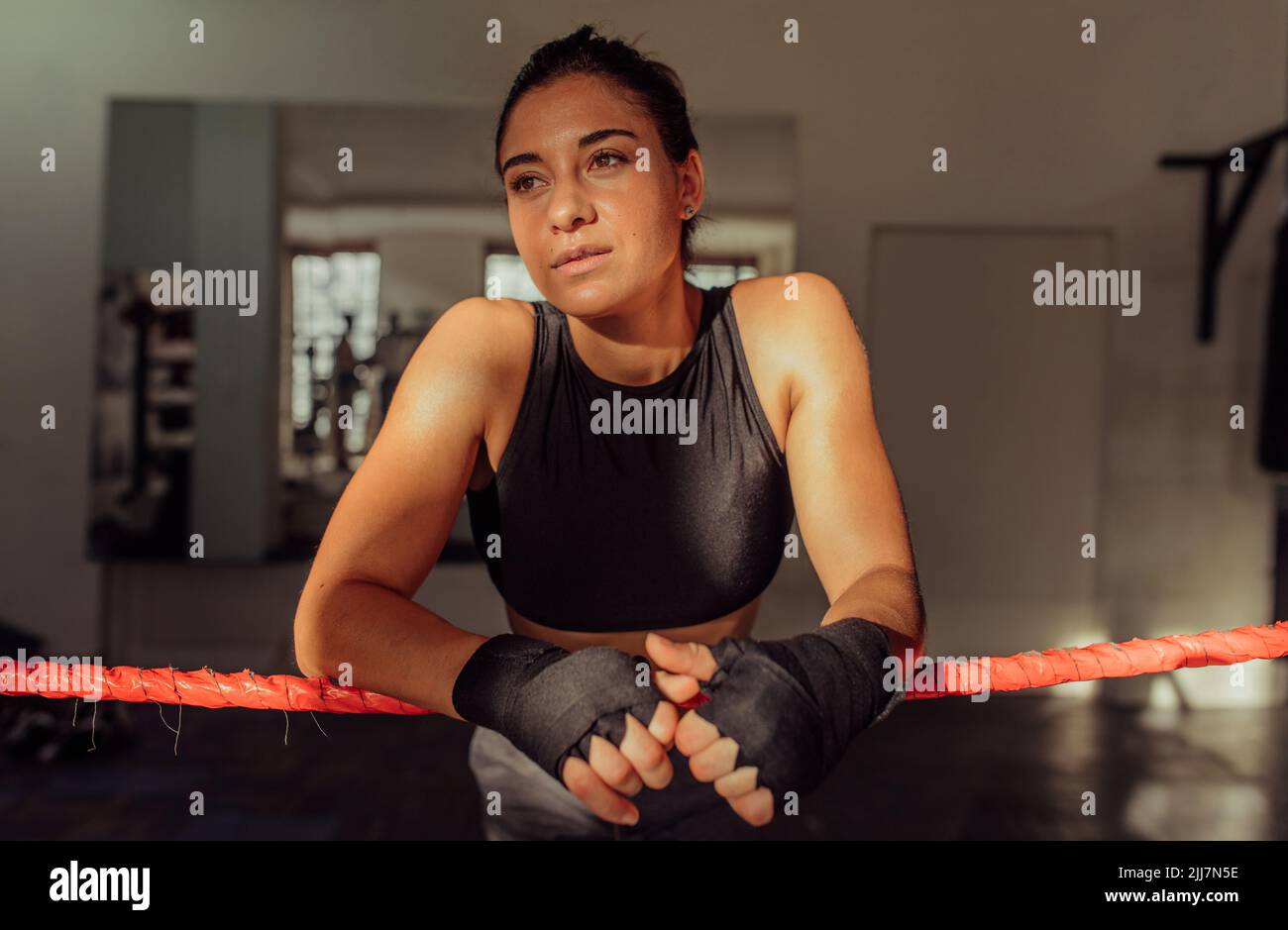 Young female boxer looking away thoughtfully while leaning on the ropes ...